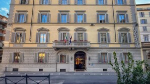 Um hotel de vários andares com estilo arquitetônico clássico, apresentando uma entrada central com varanda, diversas janelas com persianas e uma bandeira visível.