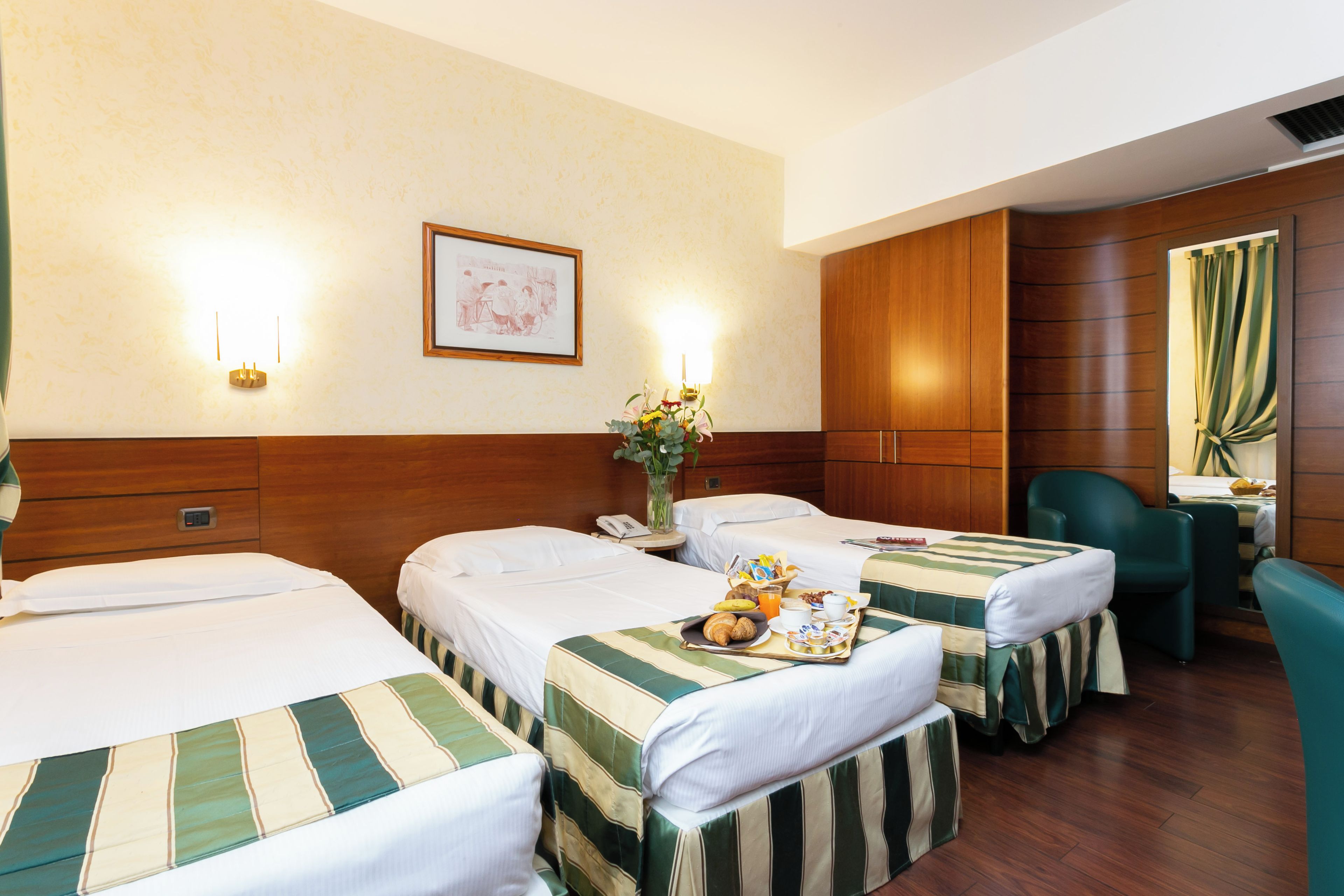 Quarto de hotel com duas camas de solteiro, área de estar com uma poltrona, escrivaninha e uma grande janela com cortinas.