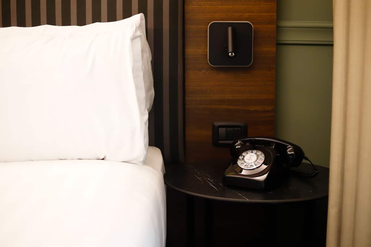 Quarto de hotel com uma cama, mesa de cabeceira com telefone rotativo e interruptor de luz na parede.