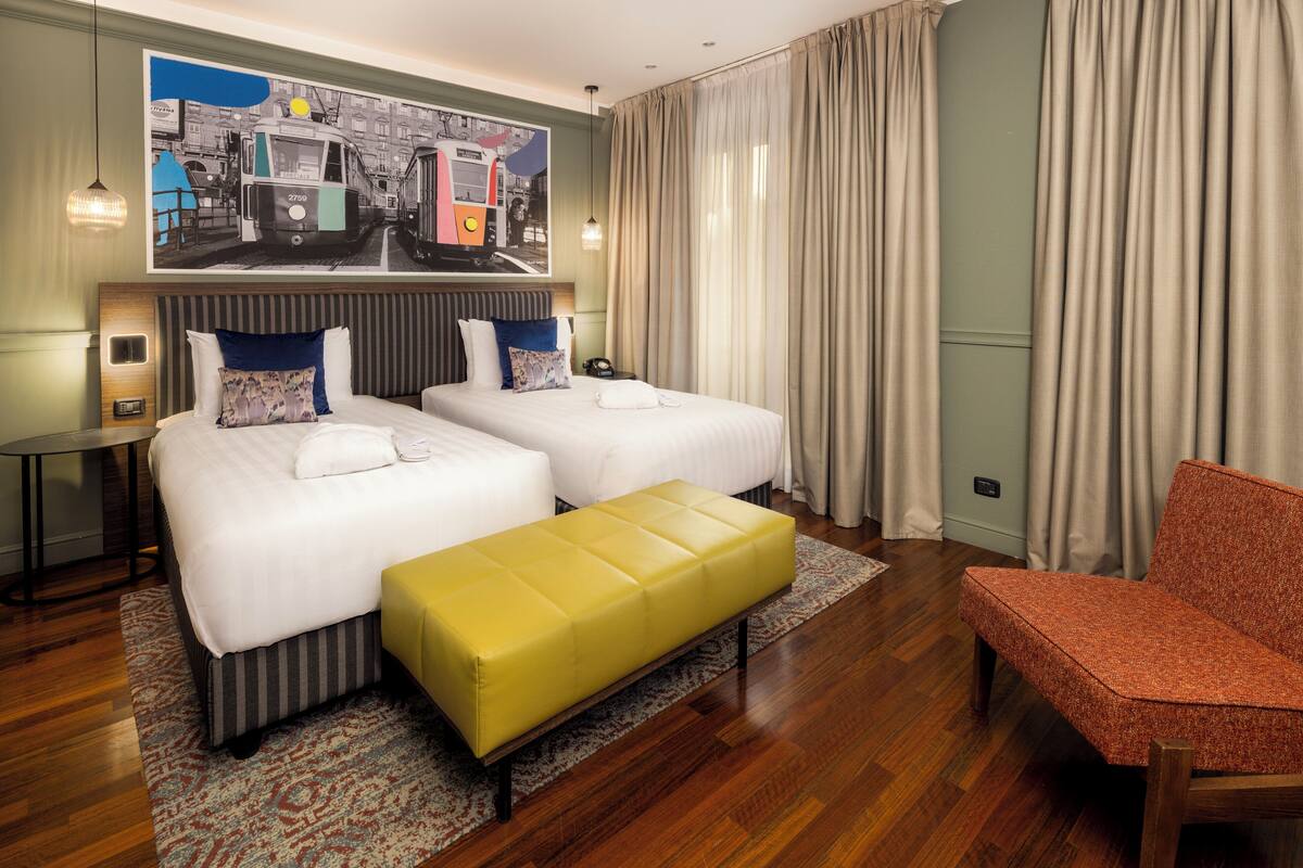 Quarto de hotel com duas camas, um banco amarelo, uma cadeira vermelha, uma pintura grande de paisagem urbana e piso de madeira.