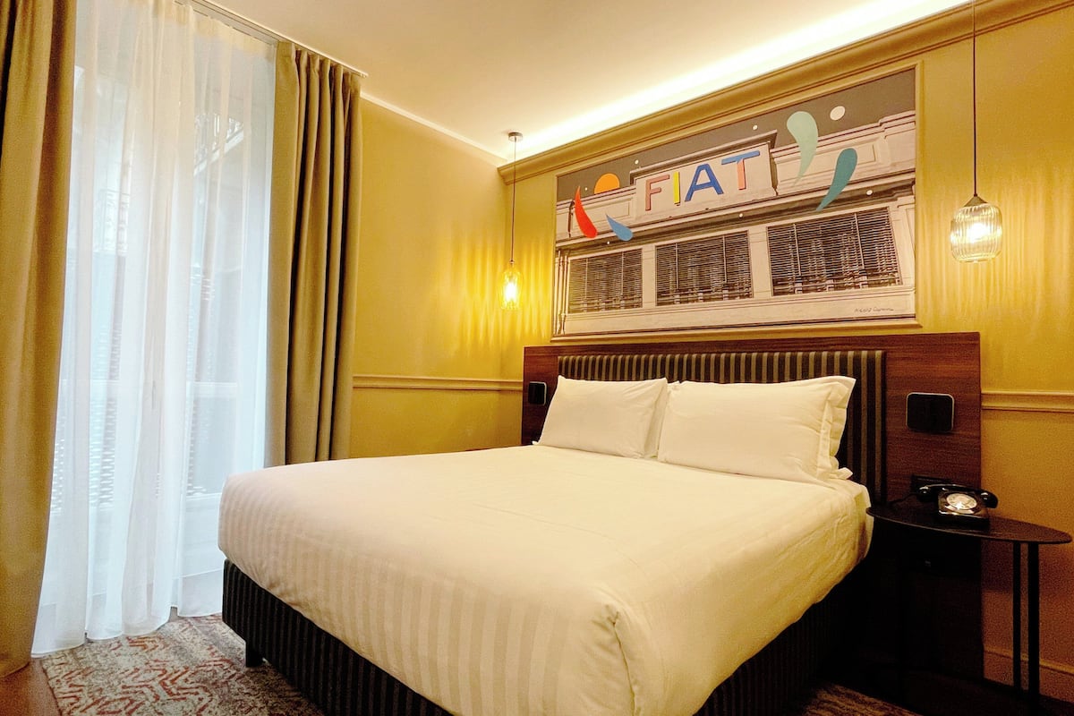 Quarto de hotel com uma cama grande, mesa de cabeceira com telefone e janela com cortinas.