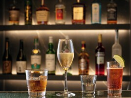 Uma área de bar com diversas garrafas de bebidas alcoólicas nas prateleiras ao fundo e três copos com bebidas diferentes no balcão.