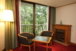 Quarto com duas cadeiras, uma mesa, um abajur, lareira e vista para árvores através de uma janela com cortinas.