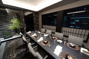 Uma sala de conferências moderna com uma mesa comprida preparada para uma reunião, vista da cidade através de grandes janelas e uma pequena planta dentro de casa.