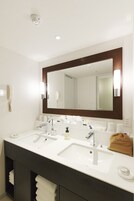 Casa de banho de hotel com duas pias, um espelho grande, um porta-toalhas e um secador de cabelo no balcão.