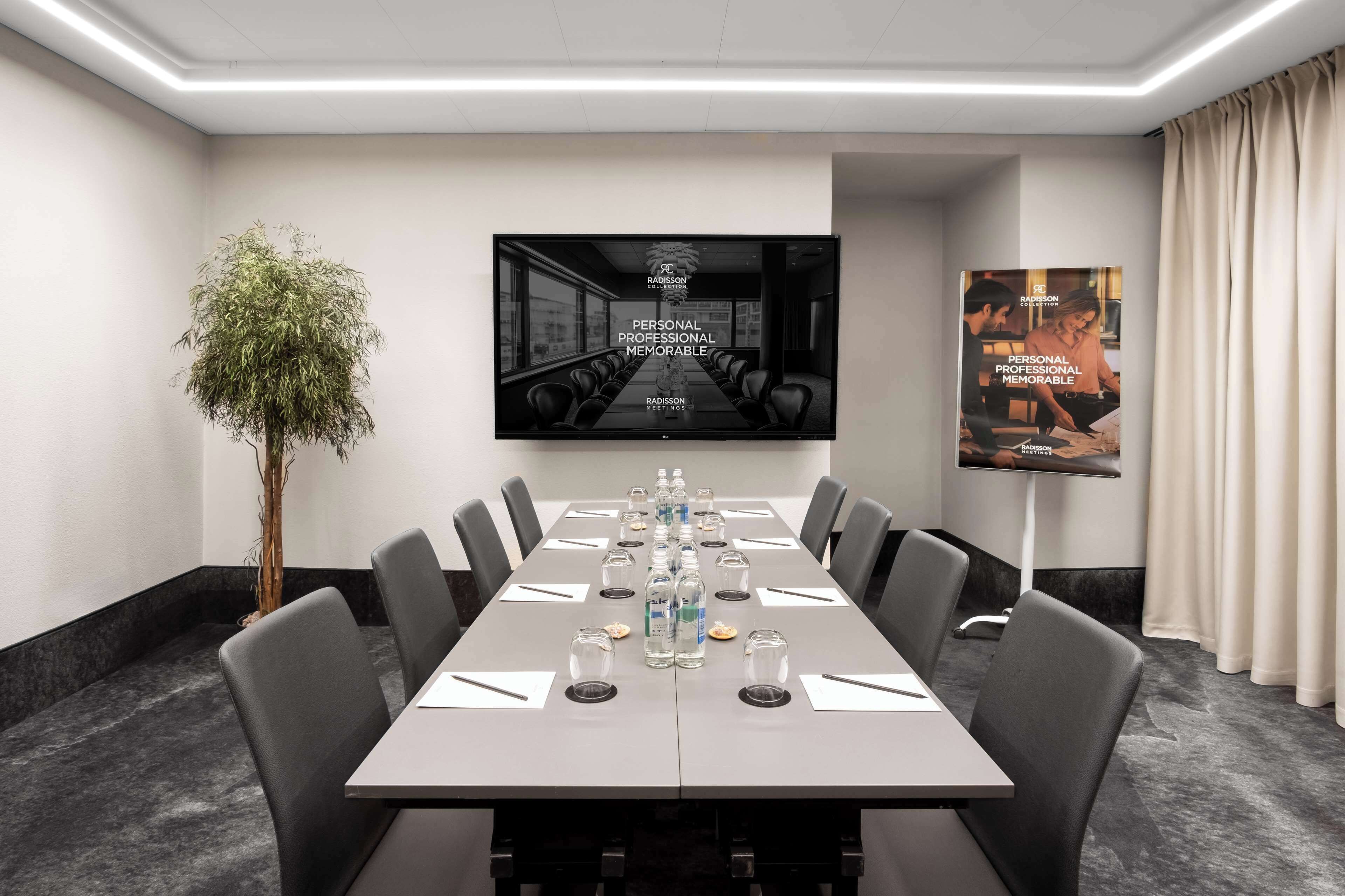 Uma sala de conferências com uma mesa comprida, cadeiras, uma planta e uma tela de televisão exibindo uma apresentação.