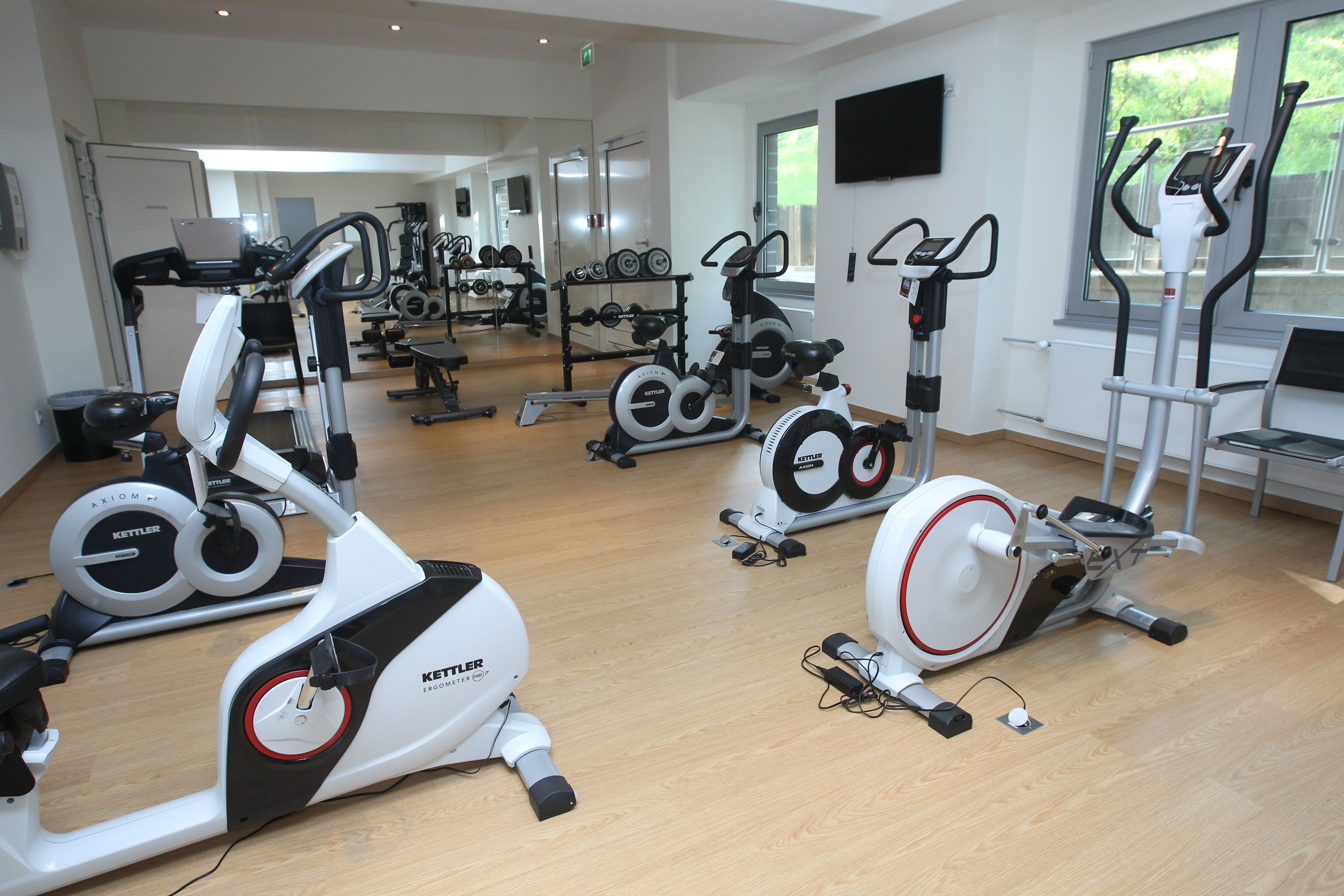 Uma academia com diversos equipamentos de exercício, incluindo esteiras e bicicletas ergométricas, dispostos numa sala ampla com grandes janelas.