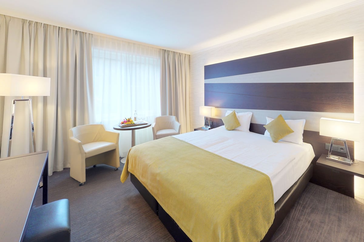Quarto de hotel com uma cama grande, duas cadeiras, uma mesinha com uma cesta de frutas e uma mesa de cabeceira com abajur.