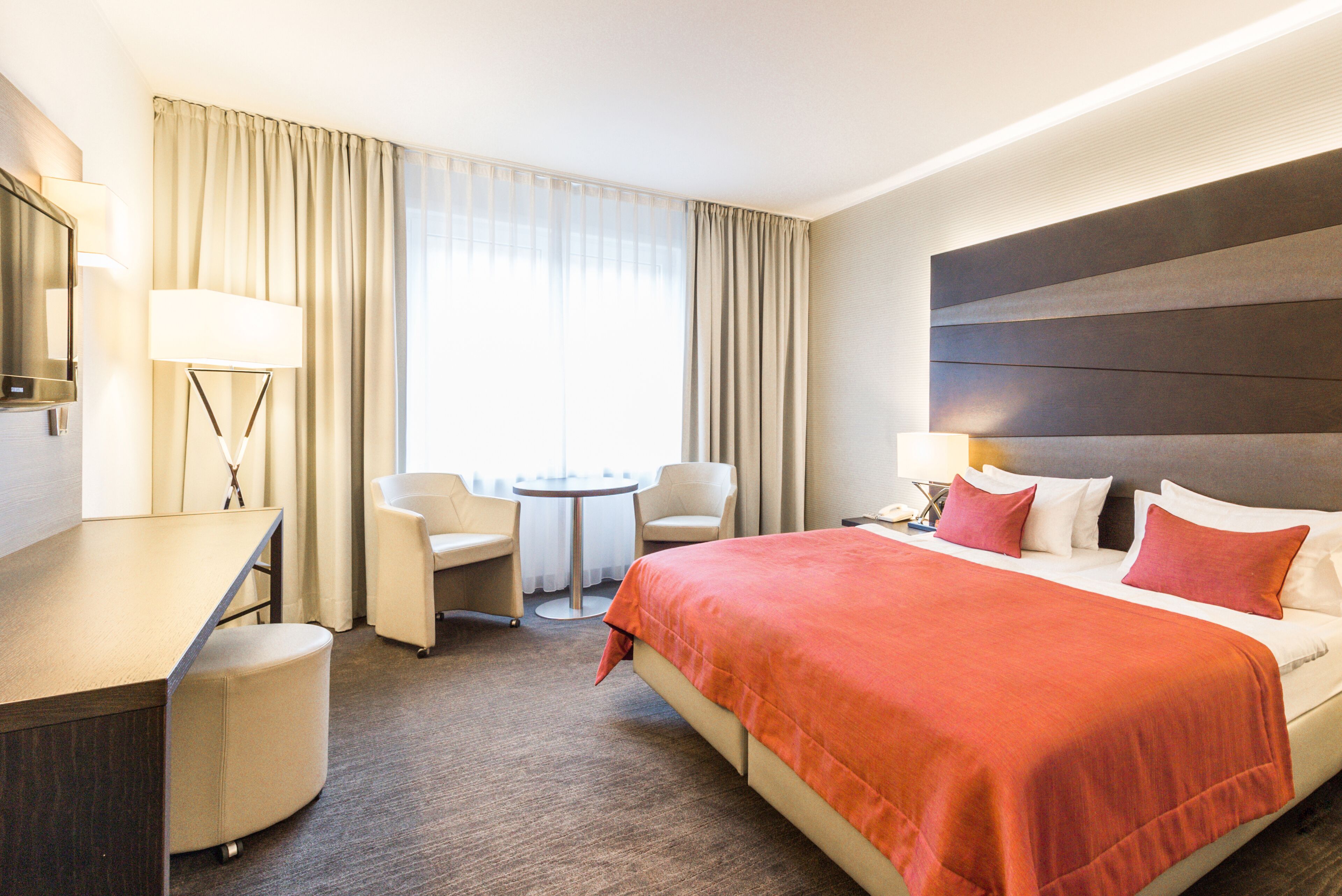 Quarto de hotel com uma cama grande, uma escrivaninha, duas cadeiras, uma mesinha e uma janela ampla com cortinas.