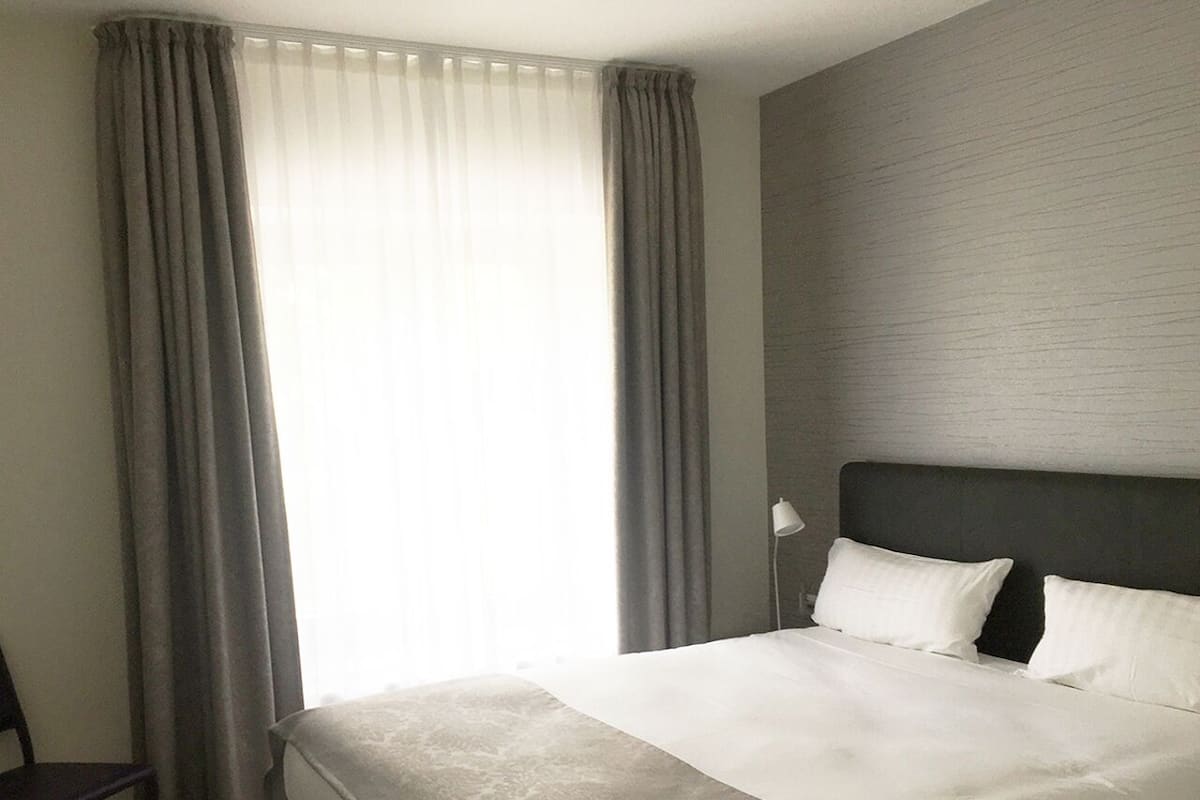 Quarto de hotel com uma cama grande, cortinas cinzas, uma cadeira e um abajur.