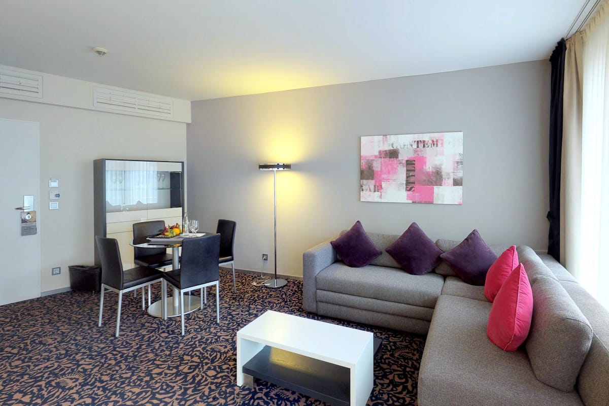 Quarto de hotel com área de jantar, sofá com almofadas, mesa de centro e abajur.