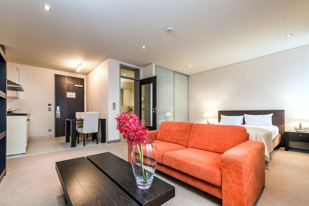 Quarto de hotel moderno com sofá, mesa de jantar, área de cozinha e cama com mesa de cabeceira.