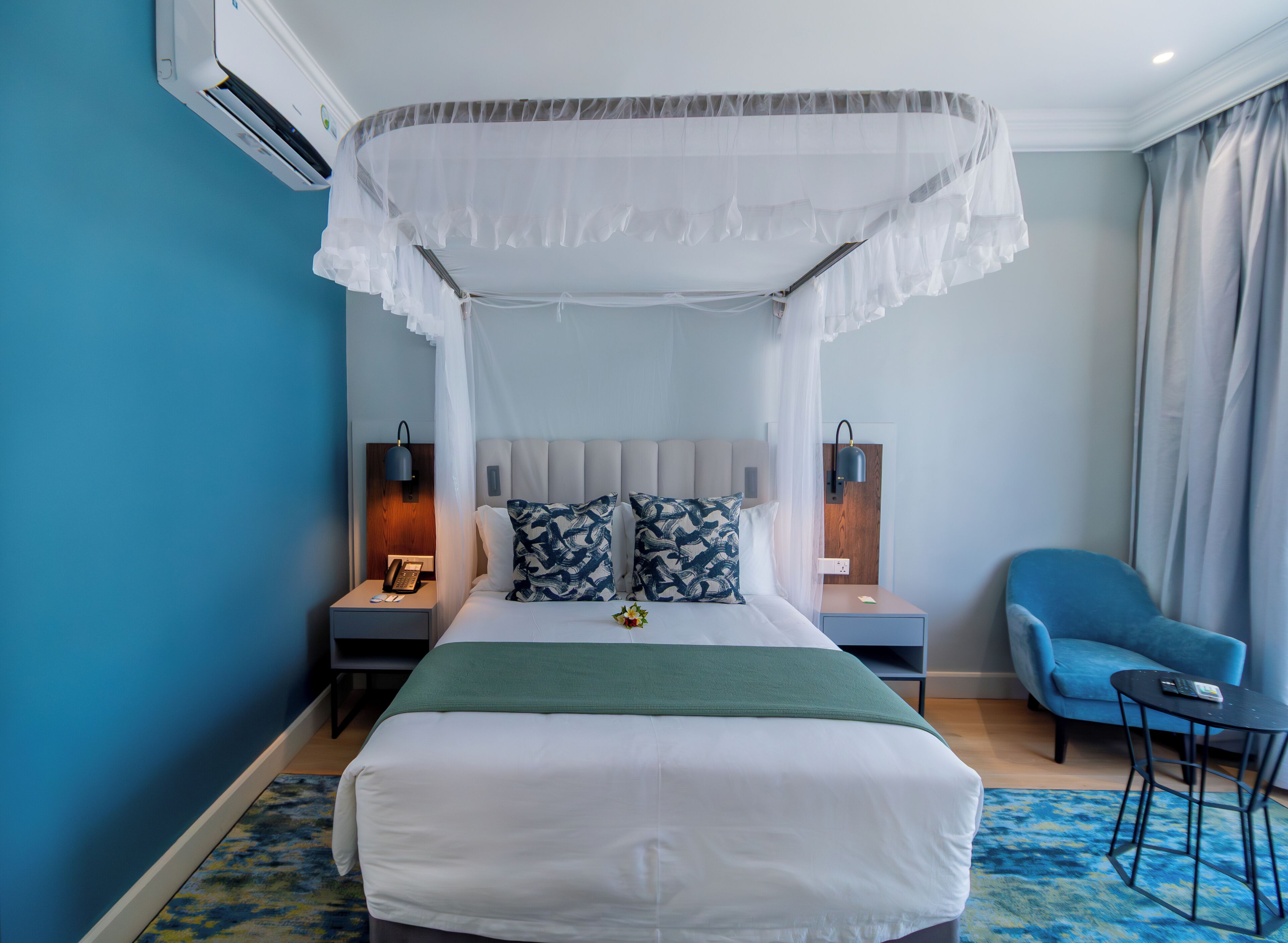 Quarto de hotel com cama de dossel, abajures, uma cadeira e uma mesinha.