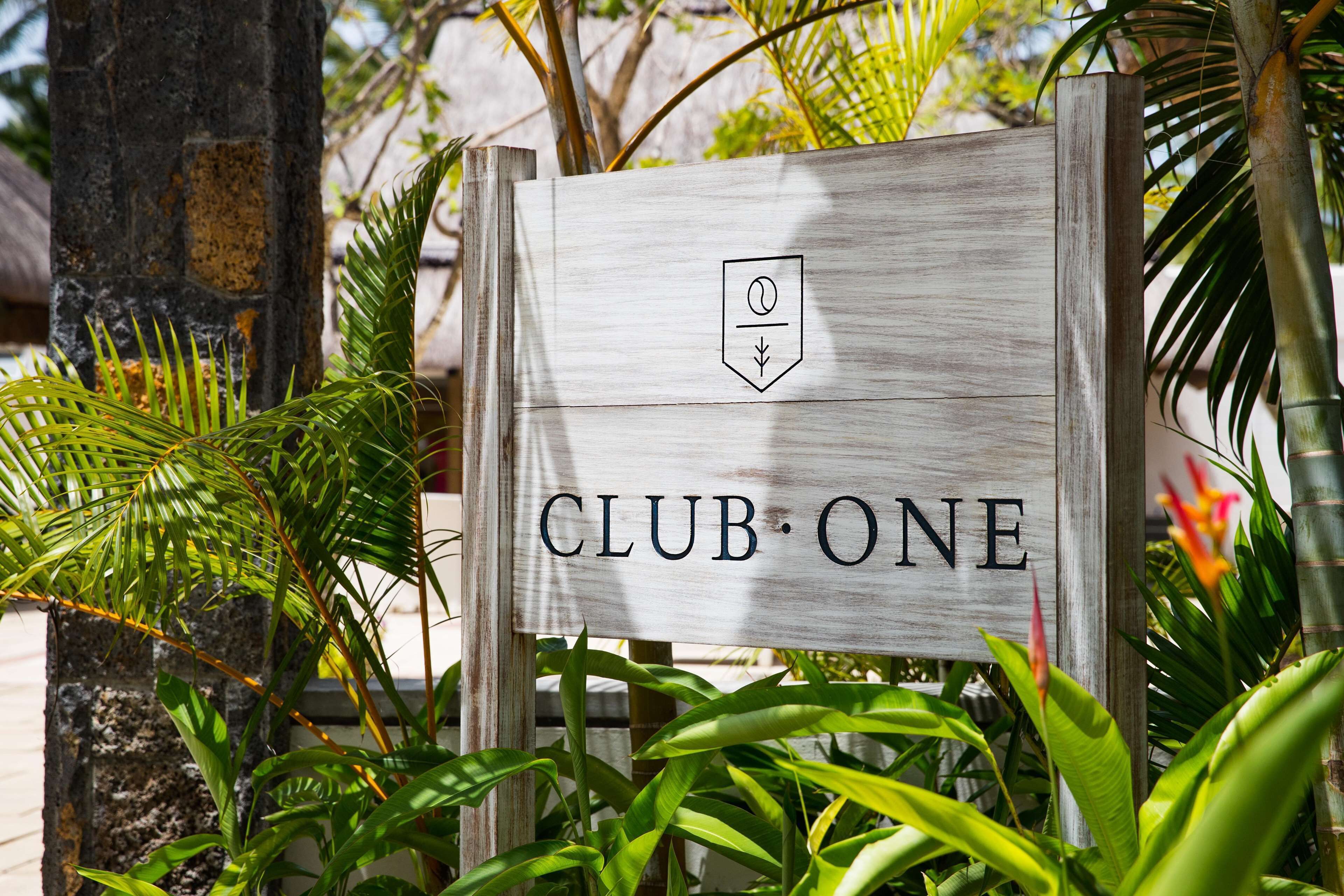 Uma placa de madeira com o texto 'CLUB ONE' rodeada por plantas tropicais.
