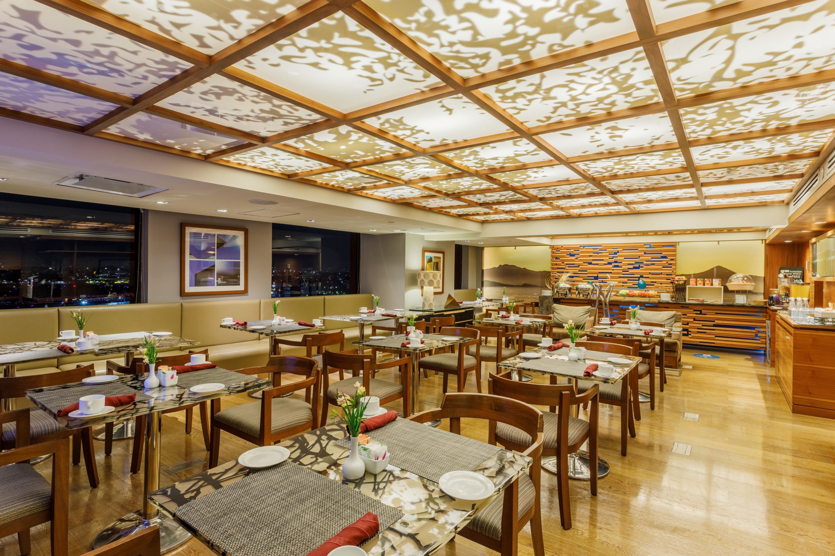 Uma sala de jantar ampla, com várias mesas e cadeiras, teto alto com design decorativo e vista para a cidade através de amplas janelas.