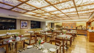 Uma sala de jantar ampla, com várias mesas e cadeiras, teto alto com design decorativo e vista para a cidade através de amplas janelas.