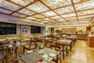 Uma sala de jantar ampla, com várias mesas e cadeiras, teto alto com design decorativo e vista para a cidade através de amplas janelas.