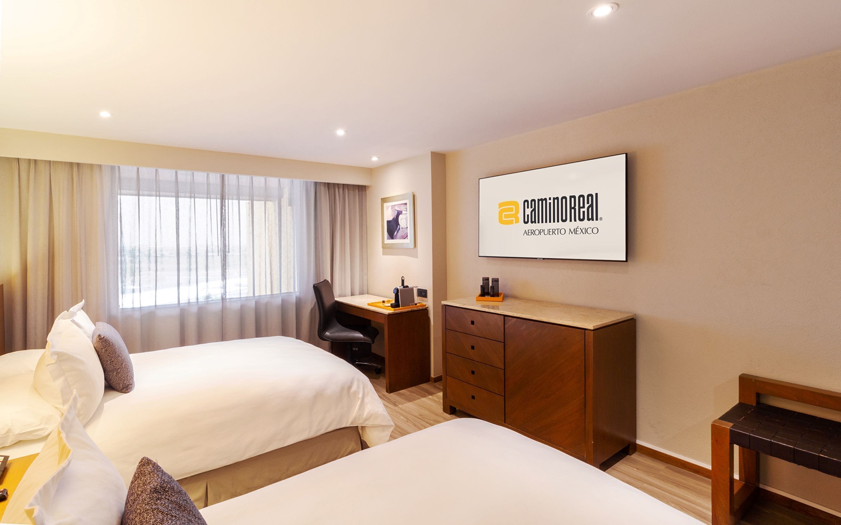 Quarto de hotel com duas camas, uma escrivaninha, uma cadeira, cômoda e uma janela grande com cortinas.