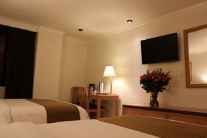 Quarto de hotel com duas camas, uma escrivaninha com abajur, televisão e um vaso com flores.