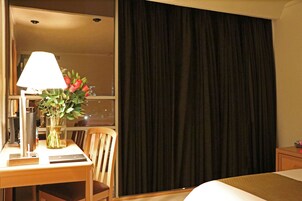 Quarto de hotel com uma cama, uma escrivaninha com luminária, uma cadeira e uma janela com vista.