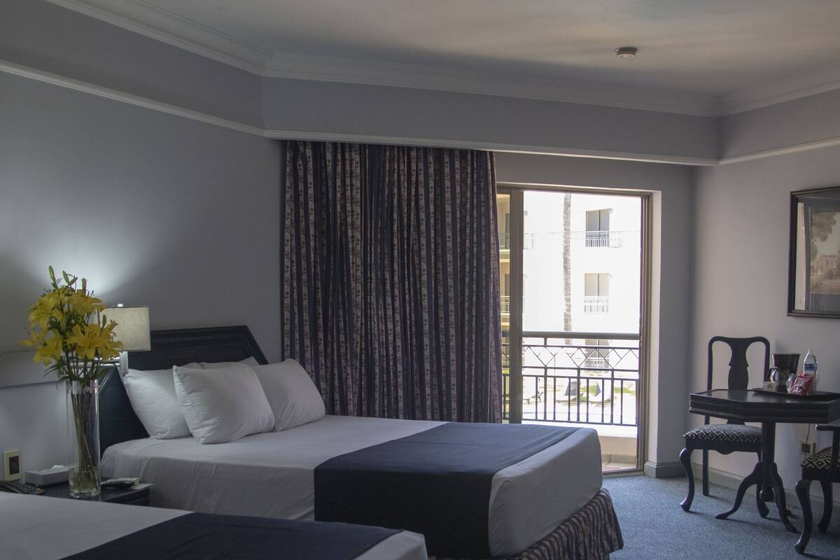 Quarto de hotel com duas camas, uma escrivaninha, uma cadeira, um vaso com flores e uma janela com cortinas.