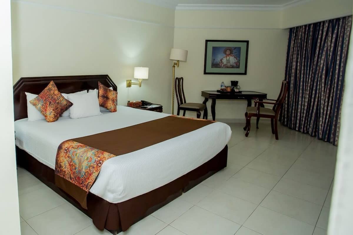 Quarto de hotel com uma cama grande, uma escrivaninha, uma cadeira, um abajur e um quadro na parede.