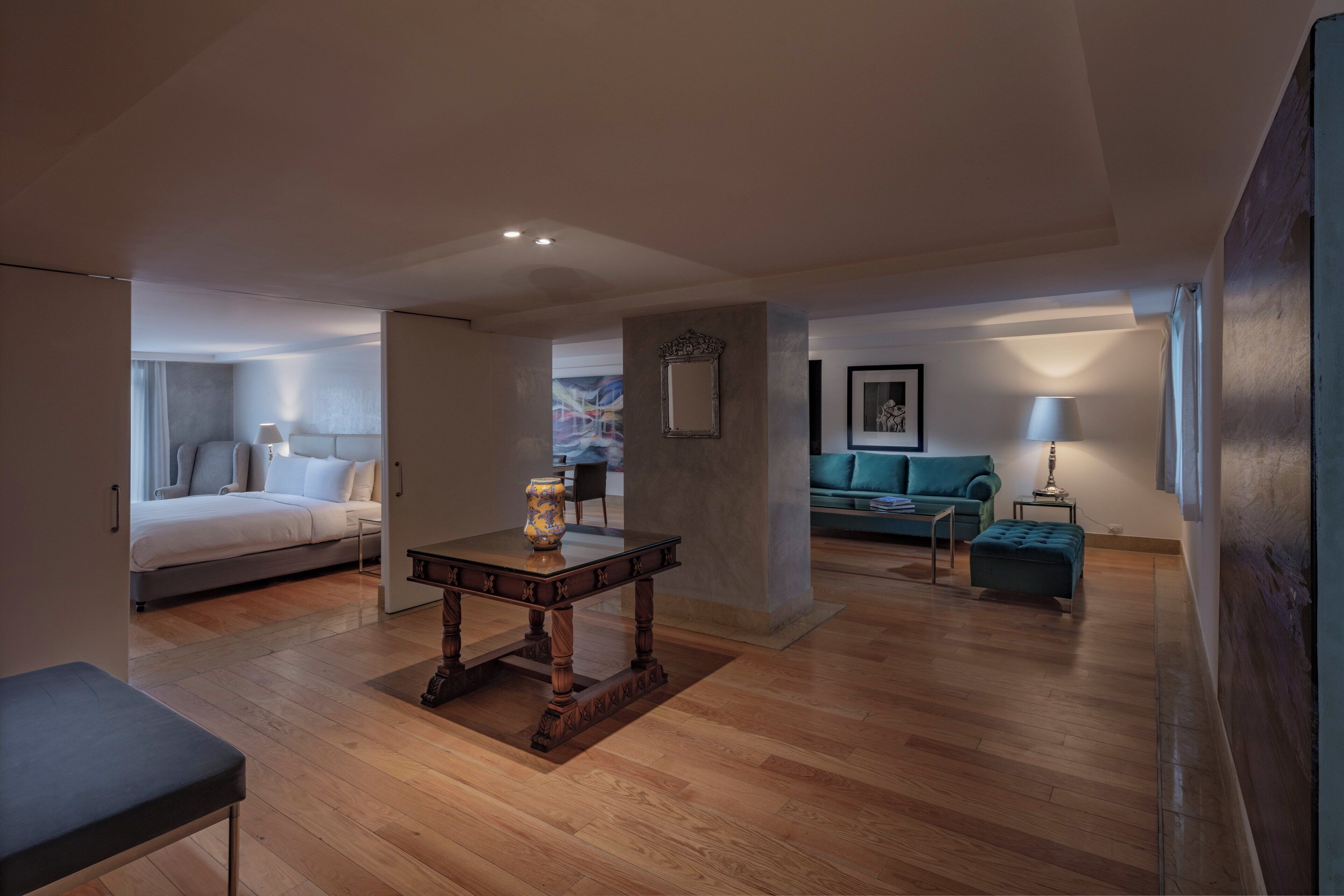 Quarto de hotel moderno com piso de madeira, cama, uma pequena mesa com um vaso, sofá e abajur.