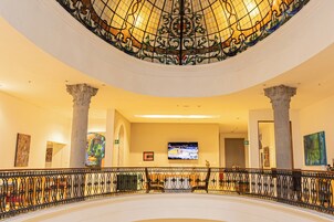 Um hall espaçoso com teto alto, grades de ferro ornamentadas e uma grande janela de vitrais.