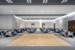 Uma sala de conferências ampla com várias mesas e cadeiras, piso coberto de carpete e um palco de madeira no centro.