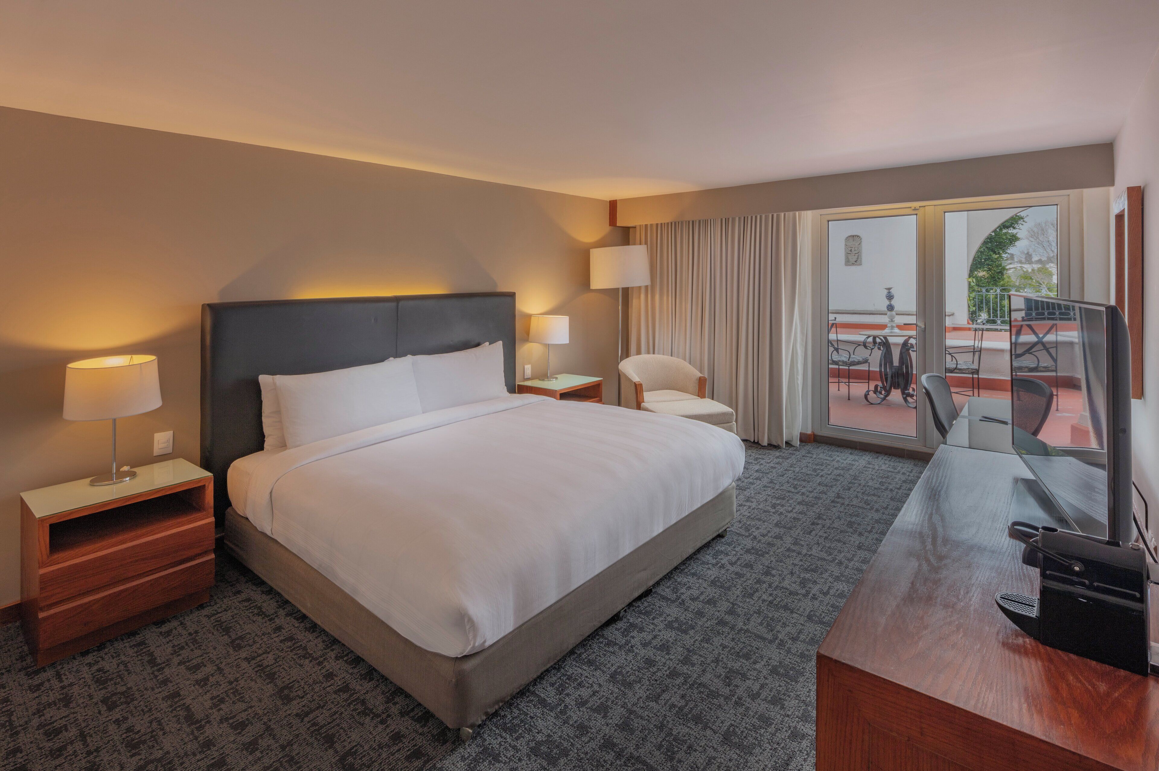 Quarto de hotel com uma cama grande, duas mesas de cabeceira com luminárias, uma escrivaninha com cadeira e vista para a varanda externa.