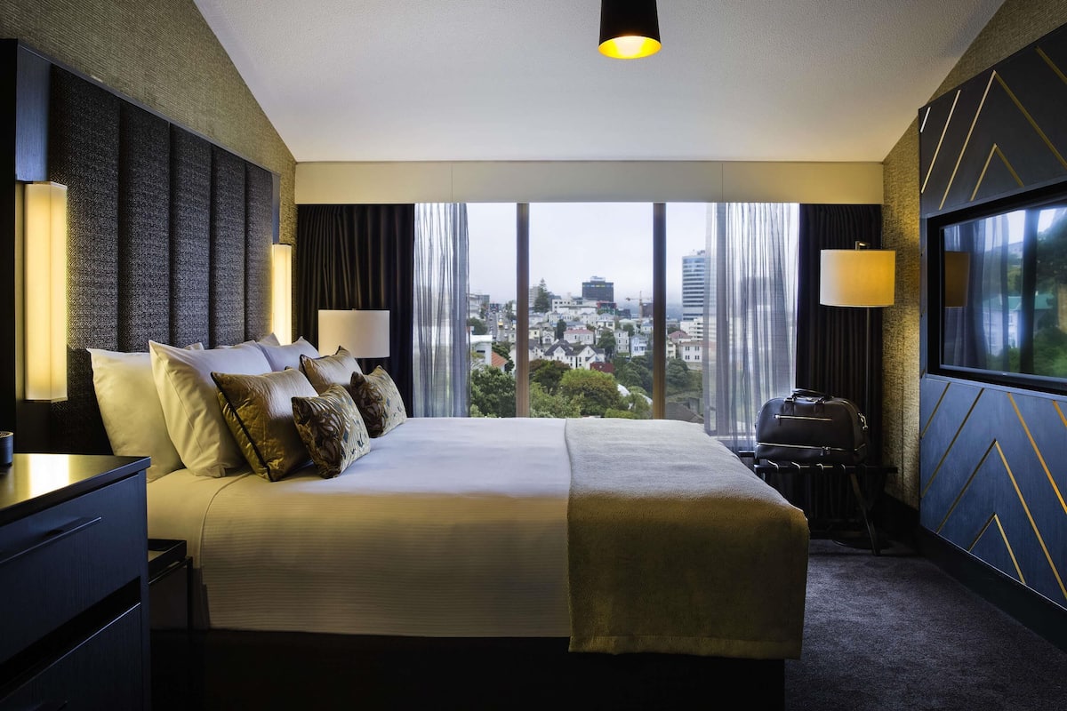 Quarto de hotel com uma cama grande, duas luminárias de cabeceira, vista para a cidade e um lustre.