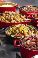 Buffet com diversos pratos em panelas vermelhas, incluindo batatas assadas, legumes grelhados e salsichas.