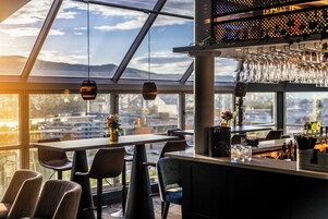 Restaurante com área de bar, mesas e vista panorâmica da cidade através de amplas janelas.