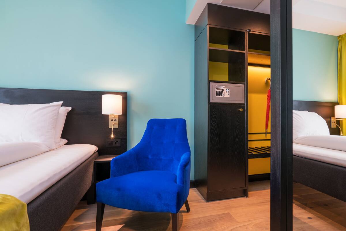 Quarto de hotel com duas camas, uma cadeira azul, um guarda-roupa e um cabideiro.