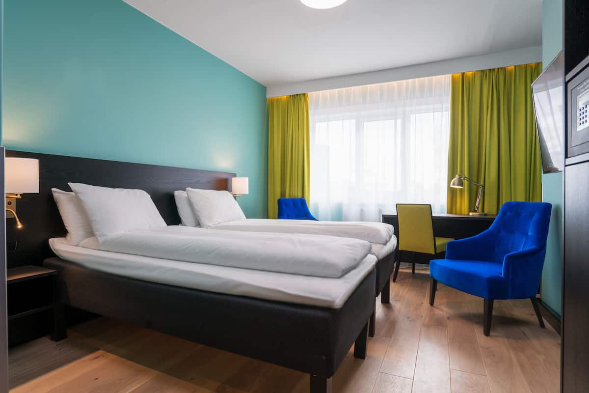 Quarto de hotel com uma cama, duas cadeiras, uma escrivaninha, televisão e janela com cortinas.
