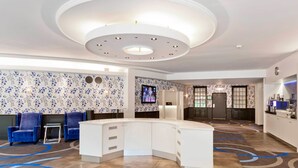 Um saguão de hotel com um balcão de recepção curvo, assentos azuis, uma tela de TV e papel de parede estampado.