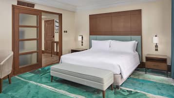 Quarto de hotel com uma cama grande, um banco, uma mesinha de cabeceira, uma luminária na parede e uma porta com painéis de vidro jateado.