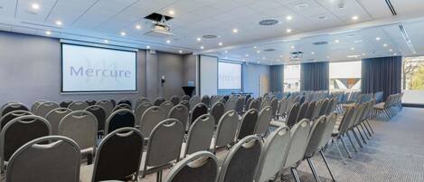 Uma sala de conferências com fileiras de cadeiras, uma tela de projetor e um janelão.