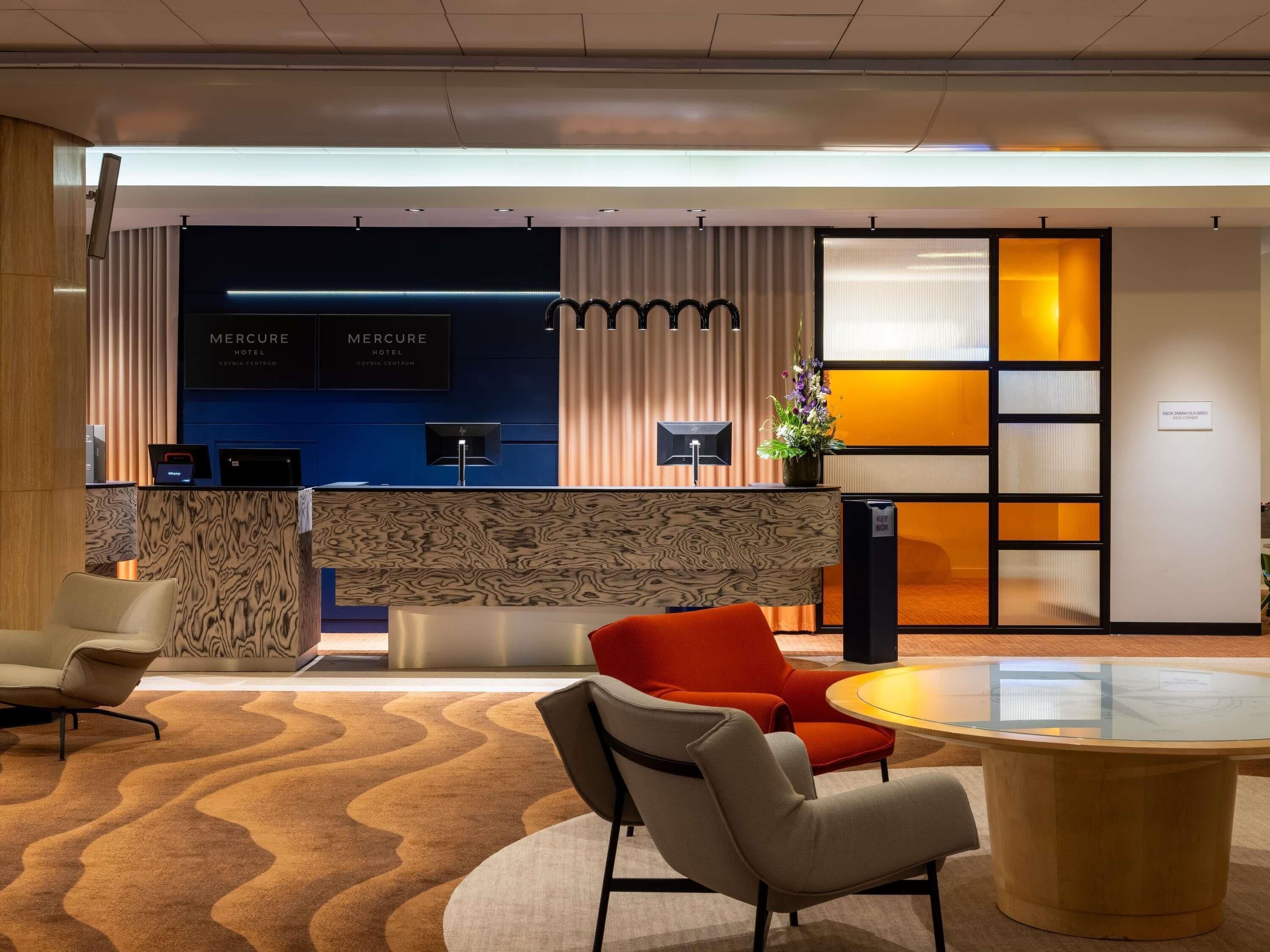 Uma área de recepção de hotel com design moderno, com balcão de atendimento, espaço para sentar e uma planta decorativa.