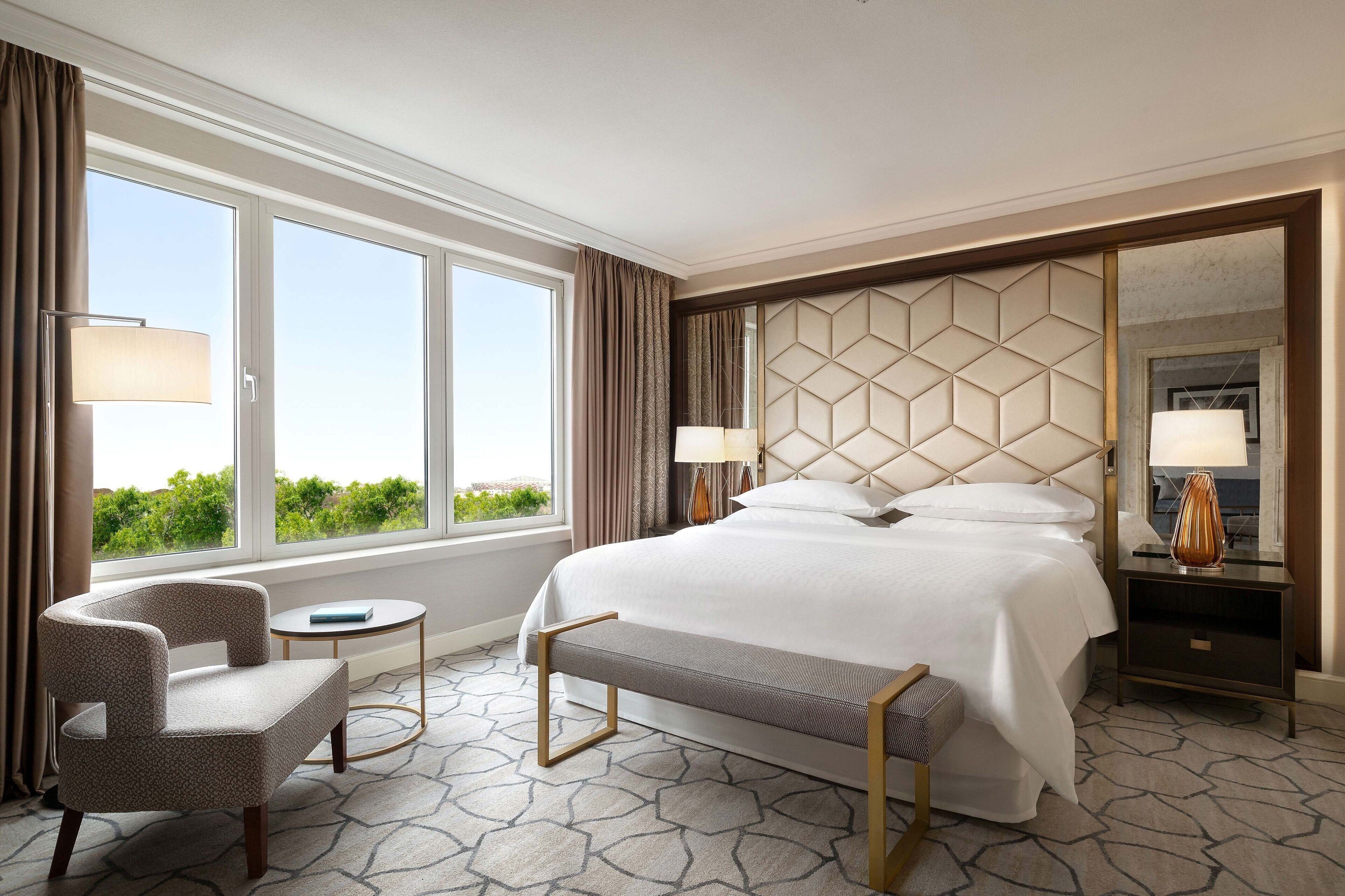 Quarto de hotel com uma cama grande, duas mesas de cabeceira com abajures, uma cadeira, uma mesinha e vista para o verde pela janela.