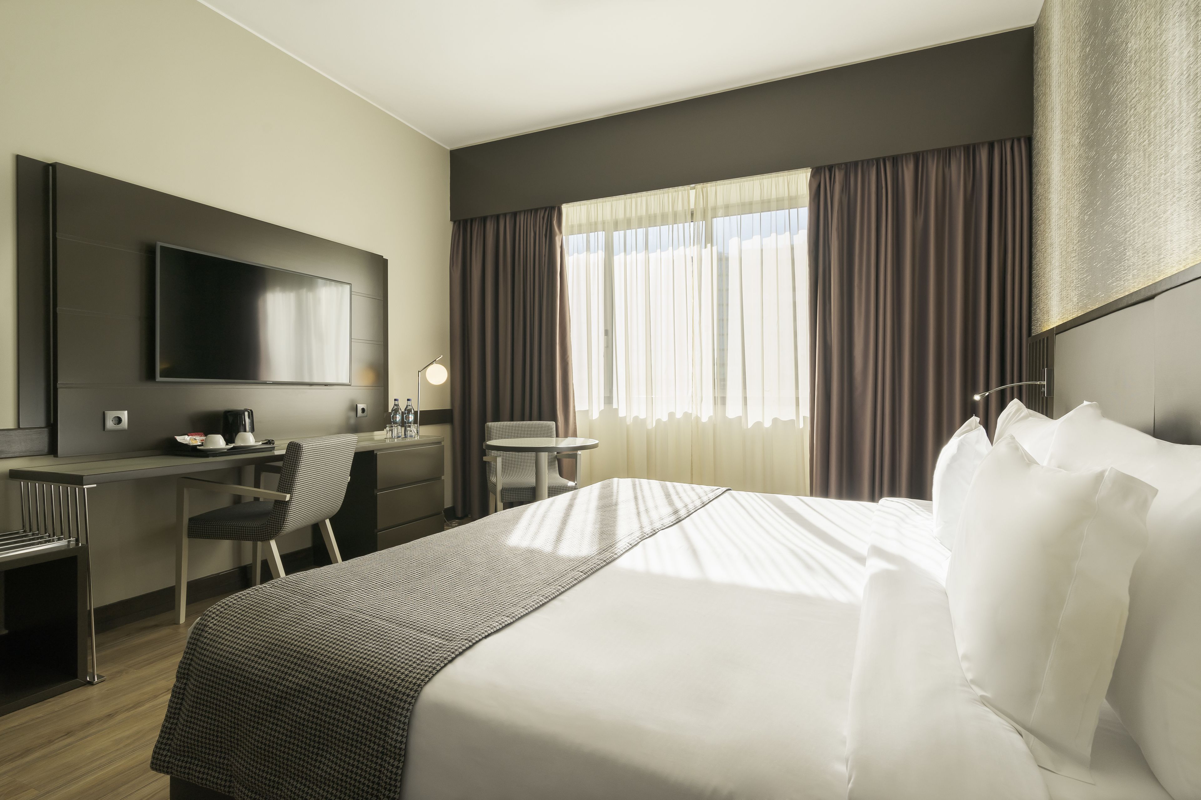Quarto de hotel com cama, escrivaninha, cadeira, televisão e cortinas.