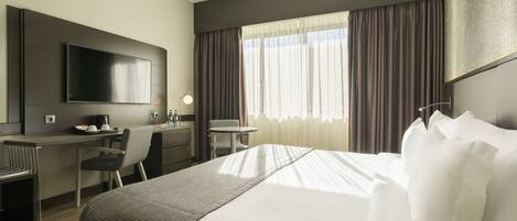 Quarto de hotel com cama, escrivaninha, cadeira, televisão e cortinas.