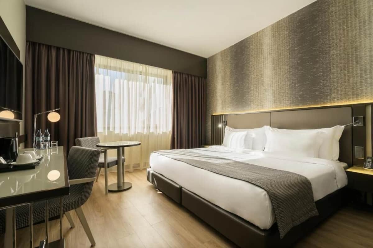 Quarto de hotel com cama, mesa de cabeceira, luminária, cadeira e vista para o exterior pela janela.