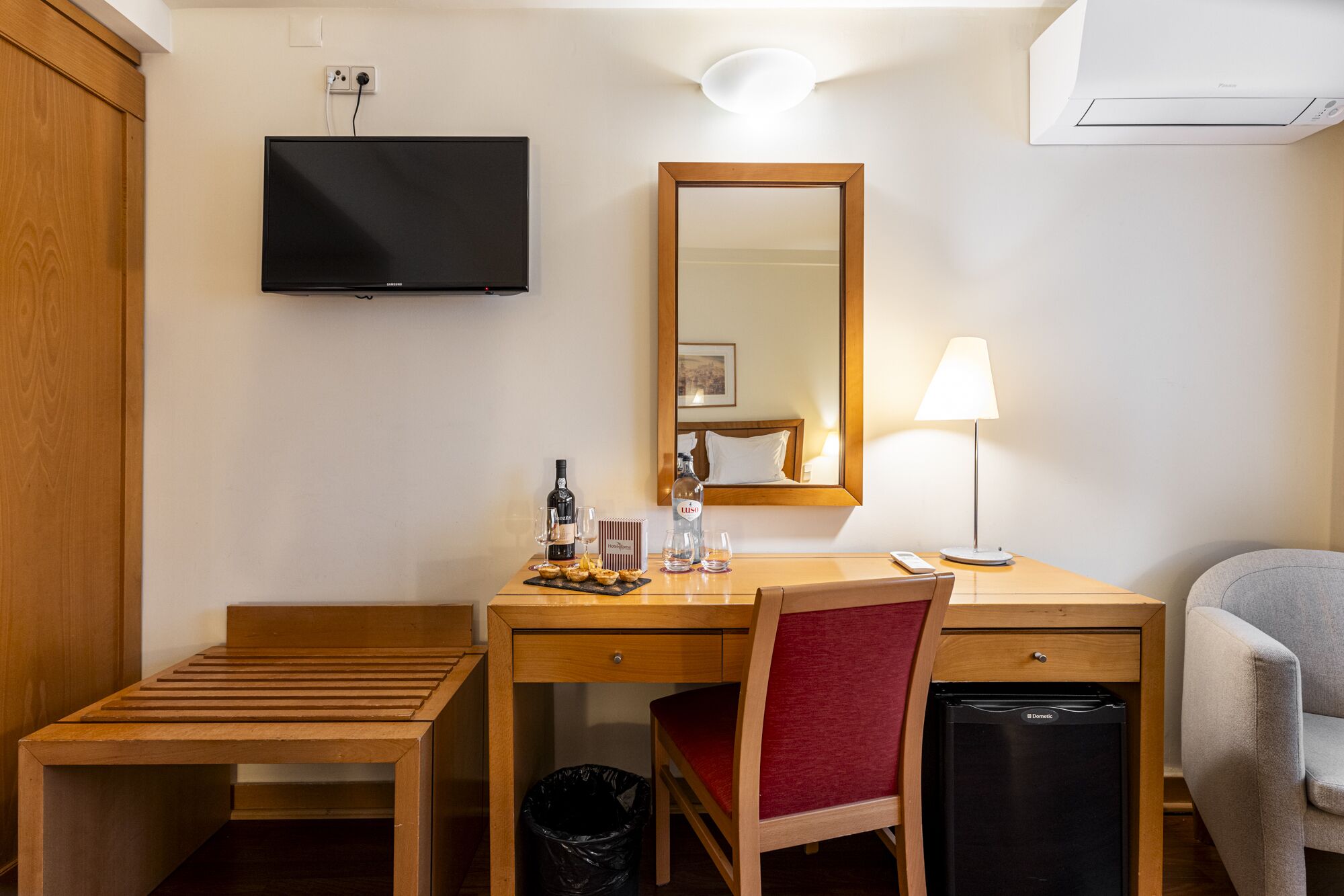 Quarto de hotel com escrivaninha de madeira, uma cadeira, um cesto de lixo pequeno, televisão, espelho, luminária e minibar.