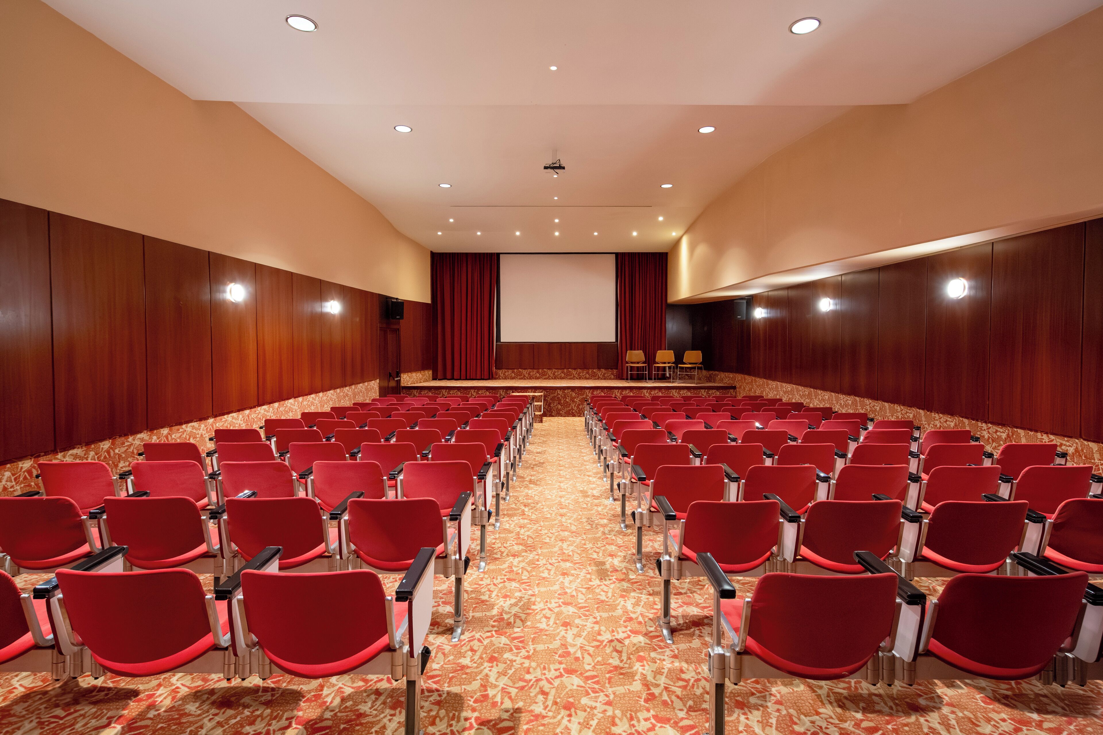 Uma sala de conferências com fileiras de cadeiras vermelhas, uma tela grande na frente e painéis de madeira nas paredes.