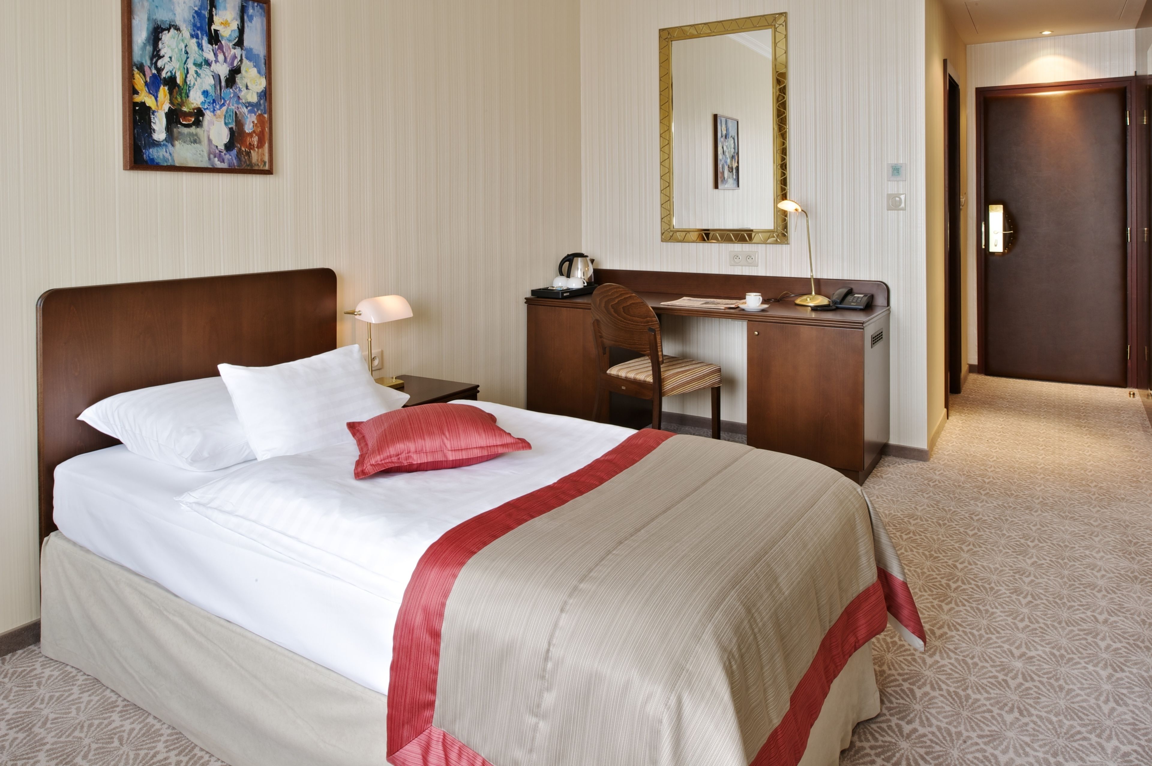 Quarto de hotel com uma cama grande, uma escrivaninha e uma cadeira. Há um quadro na parede e uma porta ao fundo.