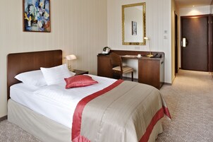Quarto de hotel com uma cama grande, uma escrivaninha e uma cadeira. Há um quadro na parede e uma porta ao fundo.