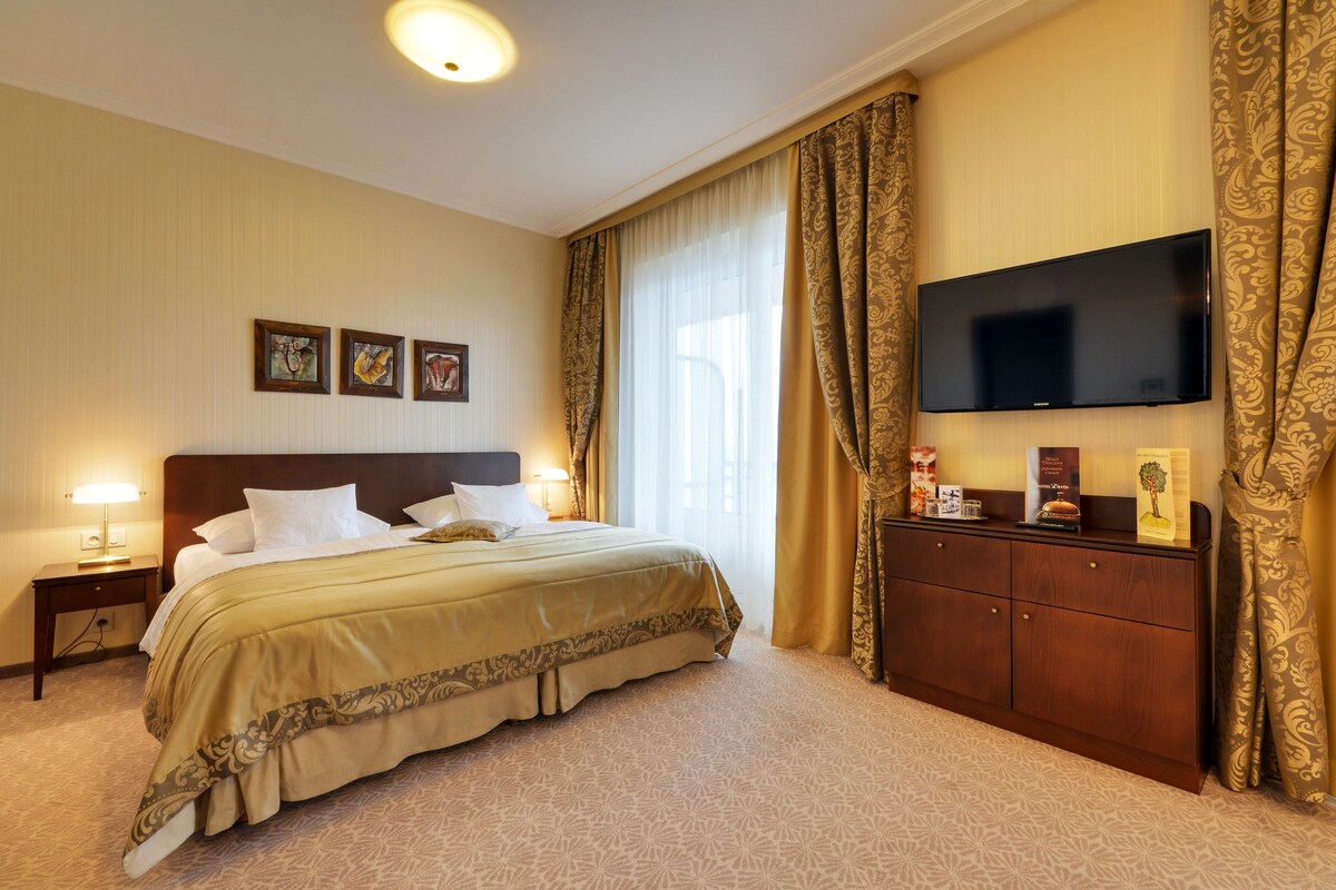 Quarto de hotel com uma cama grande, uma TV de tela plana na parede, uma mesa de cabeceira com abajur e uma cômoda de madeira com espelho.