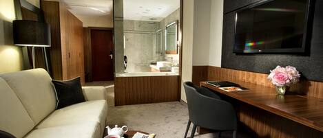 Quarto de hotel moderno com sofá, mesa de jantar com um vaso de flores, TV fixada na parede e banheiro visível através de uma porta aberta.