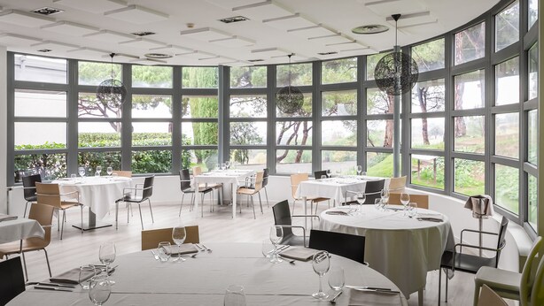 Restaurante com mesas redondas, toalhas de mesa brancas e cadeiras. Janelas amplas oferecem vista para o verde lá fora.
